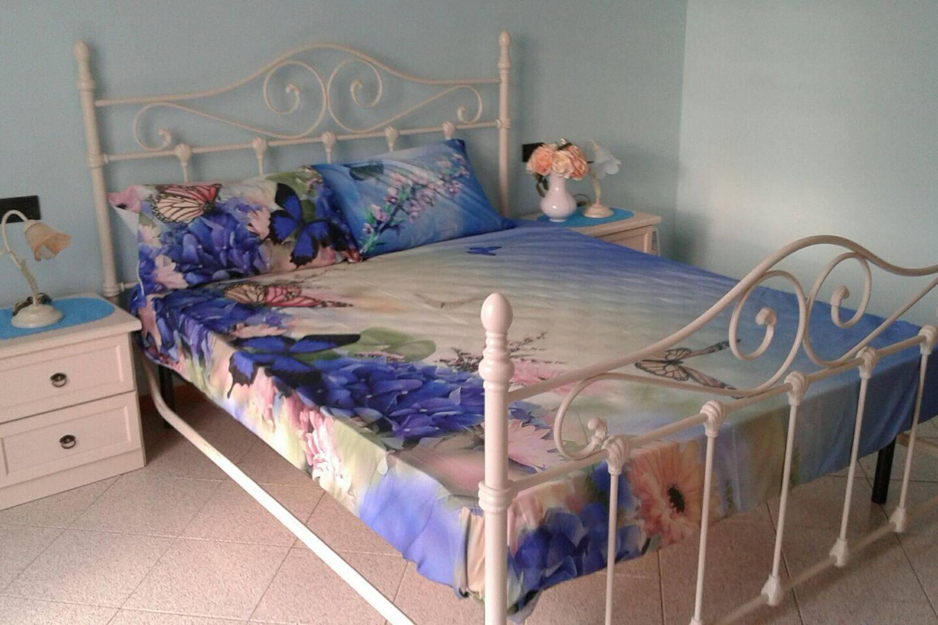 Quarto com colcha florida, estrutura de cama em metal branco, mesa de cabeceira com abajur e um vaso com flores sobre a mesa.