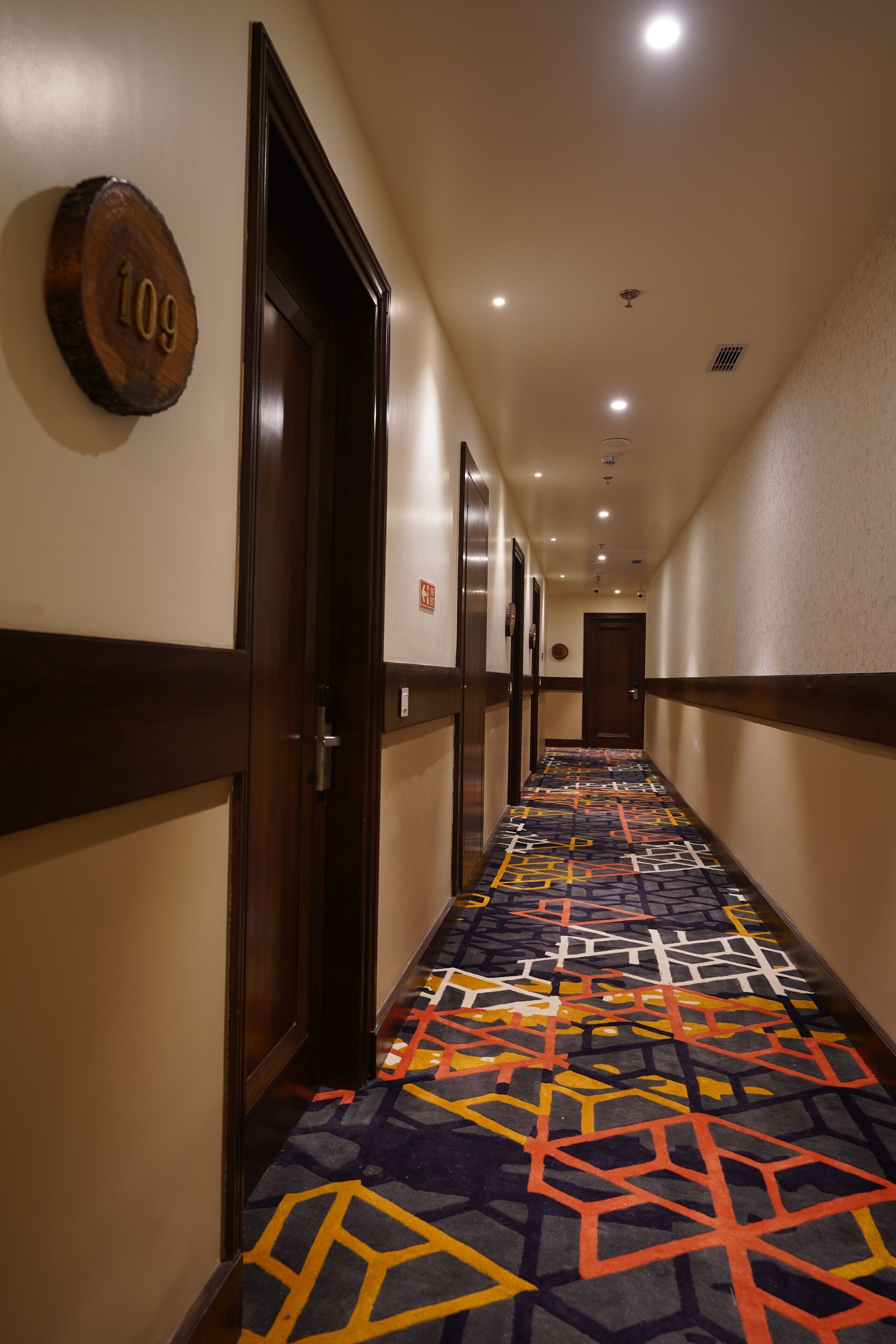 Um corredor de hotel com carpete estampado, portas de madeira e uma placa de número na parede.