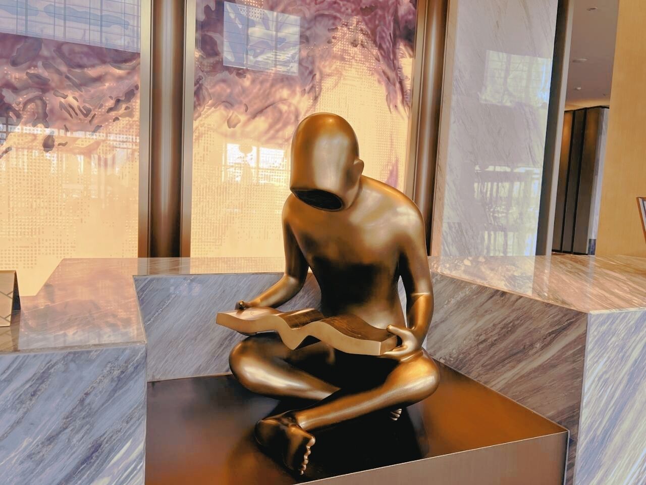 Estátua de bronze de uma figura sentada segurando um livro, colocada sobre um pedestal de mármore em um ambiente interno moderno.