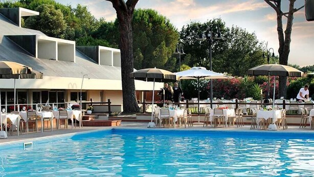 Área à beira da piscina com mesas e guarda-sóis, cercada por árvores e um edifício com design moderno.