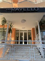Entrada de um hotel chamado RAVELLO, com porta de vidro, grades brancas e detalhes metálicos decorativos.
