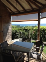 Um pátio externo com mesa e cadeiras, com vista para um jardim exuberante e o oceano.
