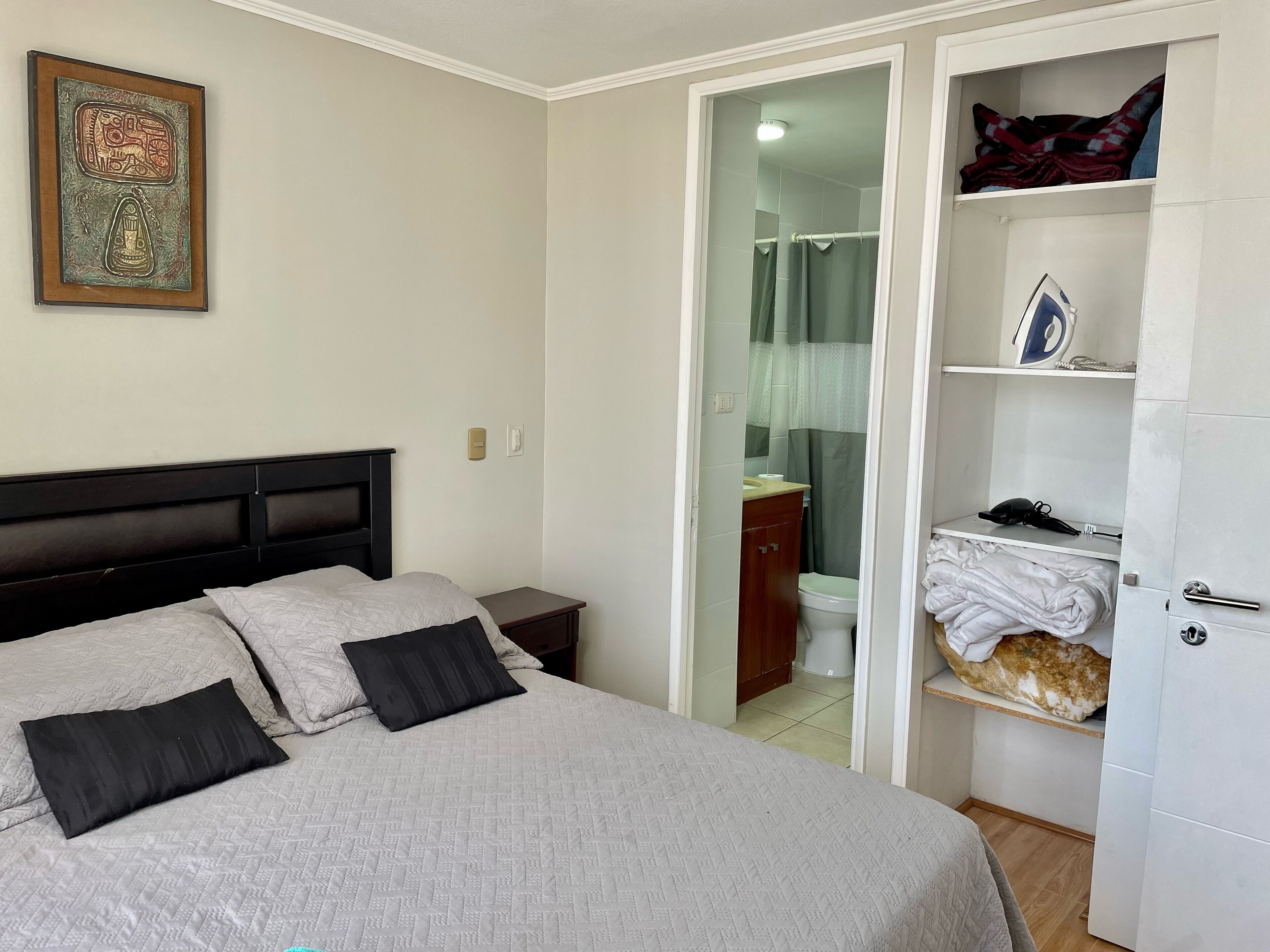Quarto com cama, mesa de cabeceira, guarda-roupa e banheiro visível por uma porta aberta.