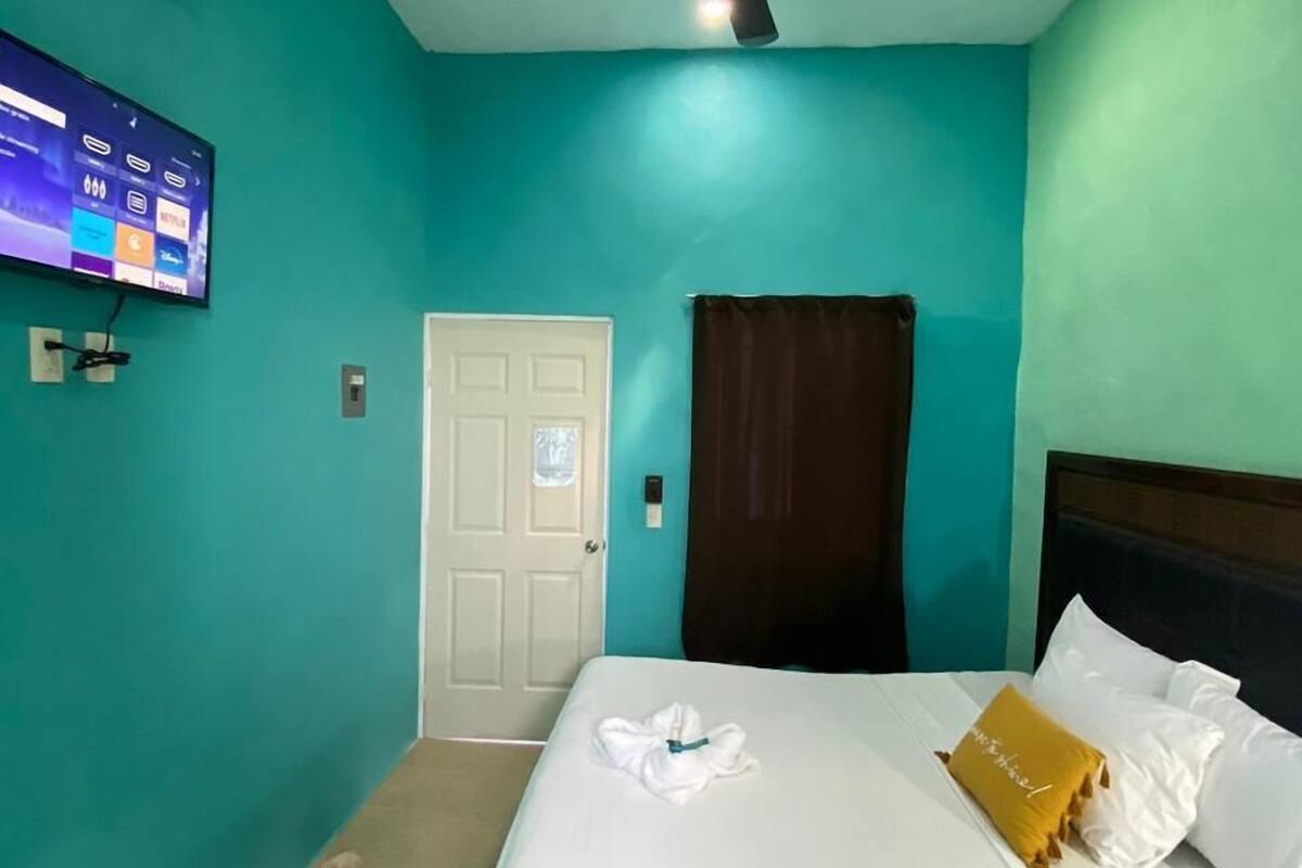 Quarto de hotel com paredes verde-água, ventilador de teto, cama com roupa de cama branca, televisão na parede e uma porta.
