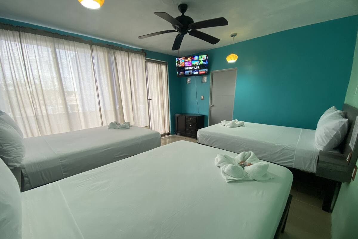 Quarto de hotel com duas camas de solteiro, um ventilador de teto, televisão na parede e uma janela com cortinas.