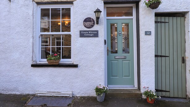 Um edifício branco com uma porta verde, uma janela com um cesto de flores e uma placa com a inscrição 'Tizzie Whizzie Cottage'.