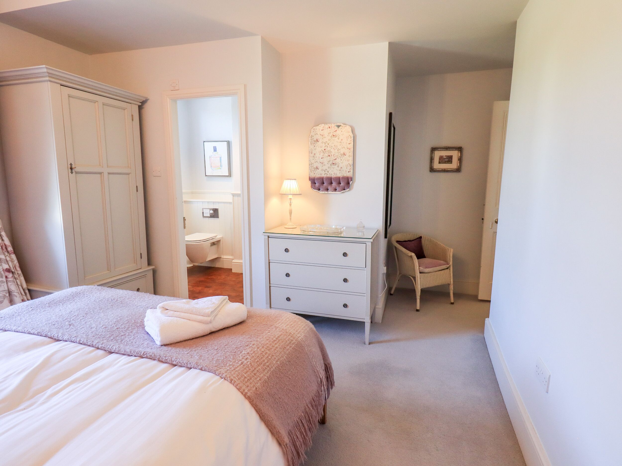 Quarto com cama, penteadeira, cadeira e banheiro visível por uma porta aberta.