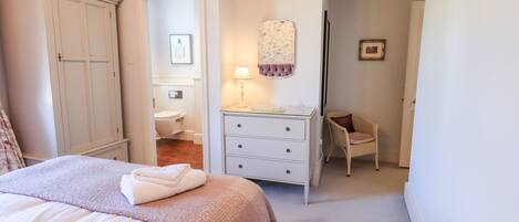 Quarto com cama, penteadeira, cadeira e banheiro visível por uma porta aberta.