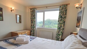 Uma cama bem arrumada com lençóis brancos, uma mesa de cabeceira de madeira com abajur e uma janela com cortinas.