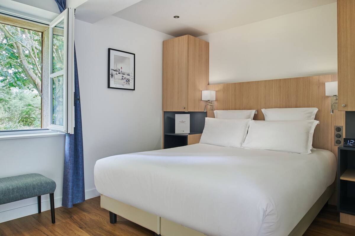 Quarto de hotel moderno com uma cama grande, uma mesa de cabeceira com abajur, um pequeno banco e uma janela com cortinas.