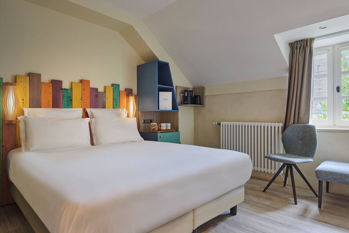 Quarto com uma cama grande, uma cadeira, uma estante com objetos e uma janela com cortinas.
