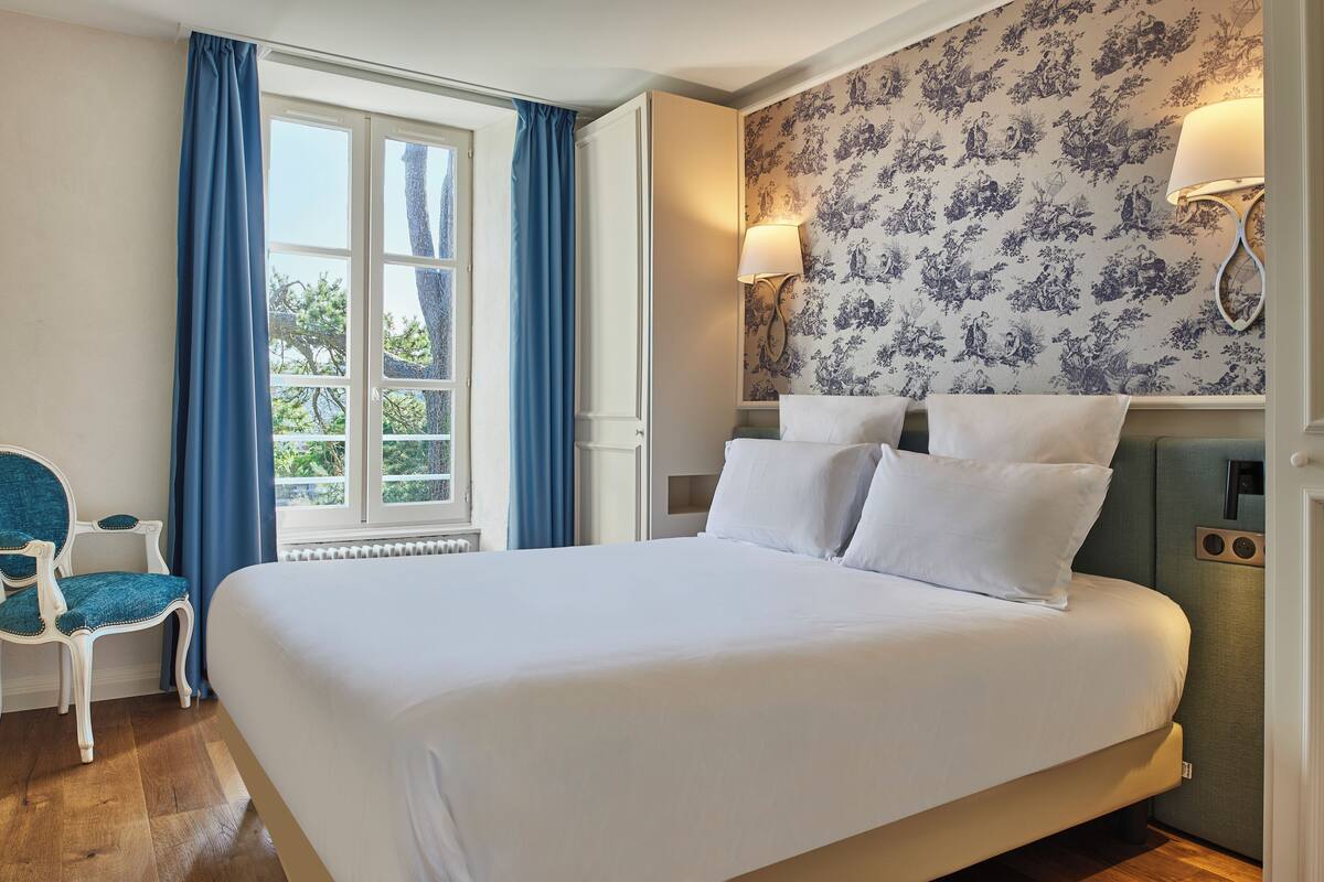Quarto com uma cama grande, uma cadeira, uma janela com cortinas e uma parede com papel de parede floral.