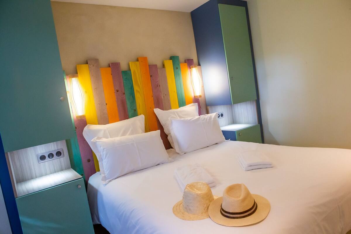 Quarto de hotel com cama, almofadas brancas, mesa de cabeceira, abajur e um chapéu sobre a cama.