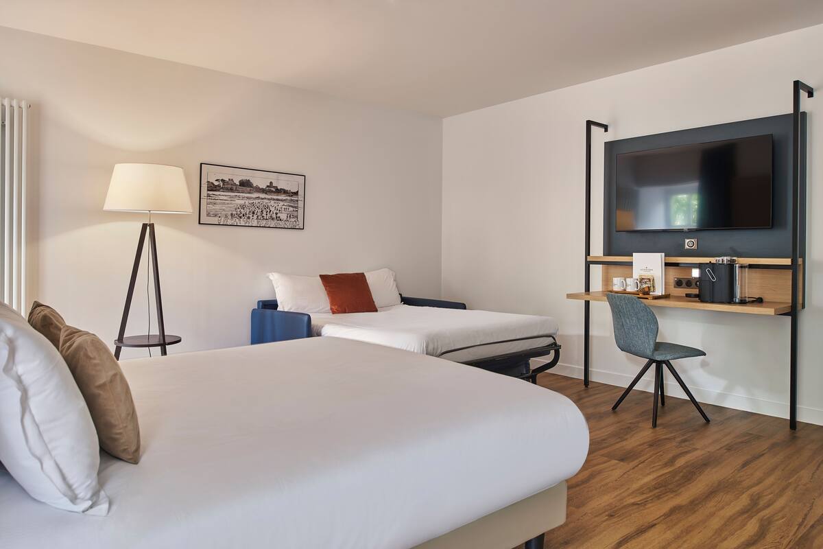 Quarto de hotel com uma cama, uma escrivaninha, uma cadeira, uma televisão, um abajur e um quadro na parede.