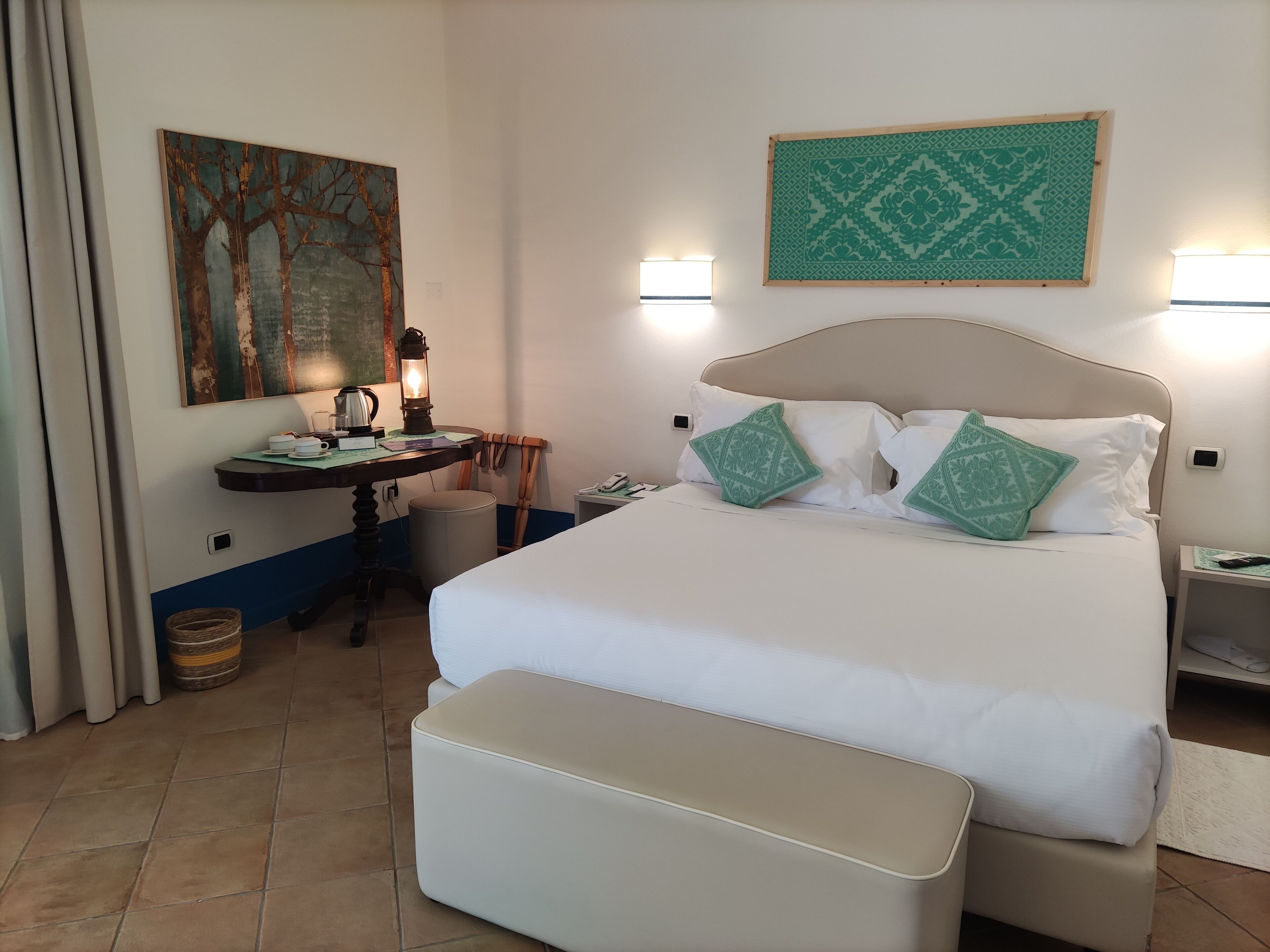 Quarto de hotel bem arrumado, com cama, mesas de cabeceira, escrivaninha com abajur, quadro na parede e tapeçaria estampada.