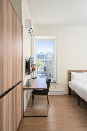 Quarto de hotel moderno com piso de madeira, cama com roupa de cama branca, escrivaninha com cadeira, televisão na parede e janela com vista para a cidade.