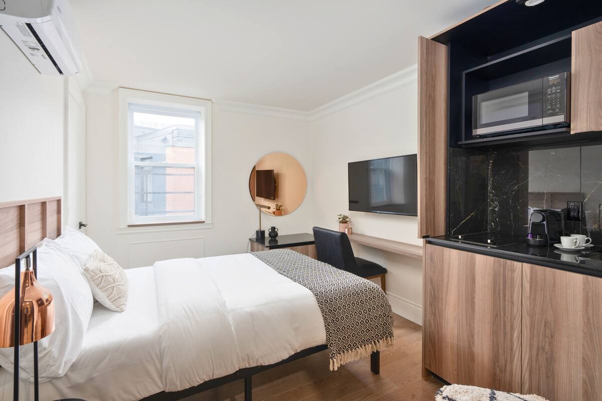 Quarto de hotel compacto com cama, escrivaninha, espelho redondo, televisão, micro-ondas e máquina de café.