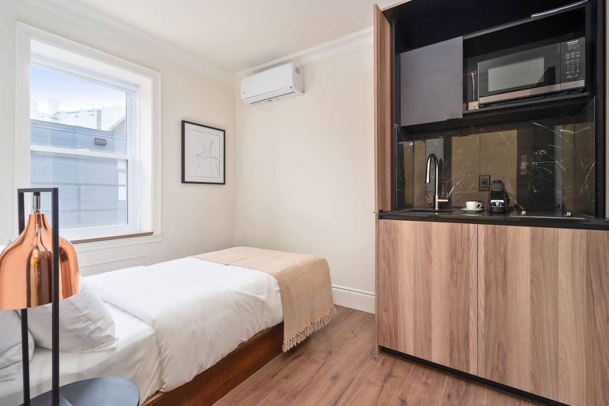 Quarto compacto com cama, pequena cozinha equipada com micro-ondas e janela com vista para edifícios.