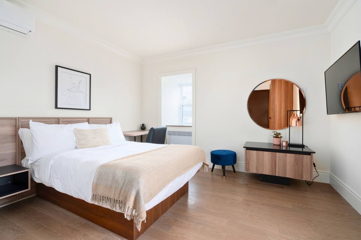 Quarto moderno com cama de madeira, uma escrivaninha, uma cadeira, um espelho redondo e um banquinho.