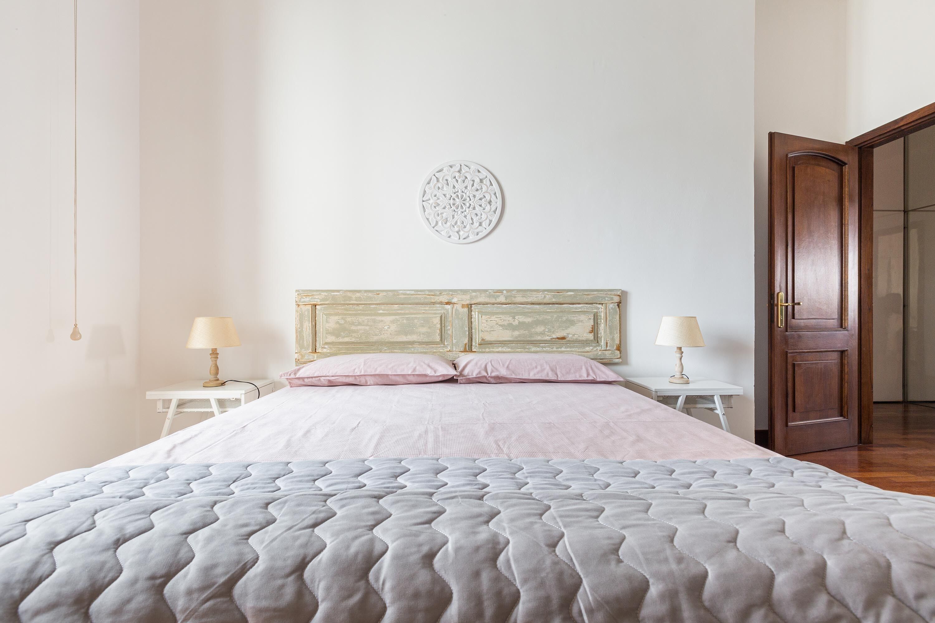 Quarto com cama, duas mesas de cabeceira, um prato decorativo fixado na parede e porta de madeira.