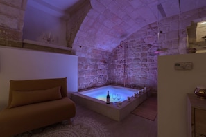 Quarto com paredes de pedra, uma banheira, um sofá e uma pequena mesa com uma garrafa e copos.