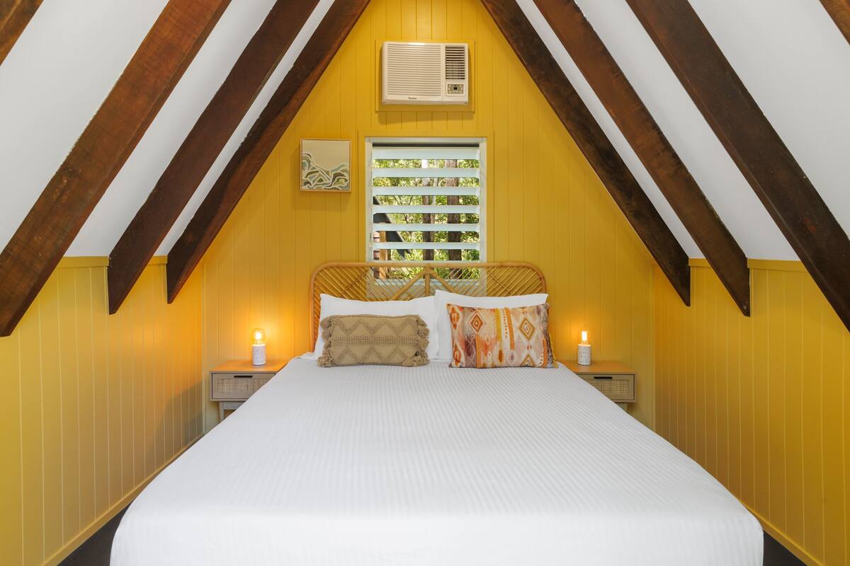 Quarto com teto inclinado, cama com roupa de cama branca, duas mesas de cabeceira com abajures e uma janela com persianas.