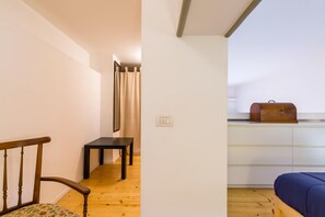 Um quarto com piso de madeira, uma cama com colcha azul, uma cadeira de madeira, uma pequena mesa e um armário com uma mala em cima.