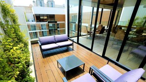 Uma área de estar moderna ao ar livre, com sofá e mesas, cercada por paredes de vidro e um deck de madeira.