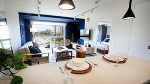 Quarto de hotel moderno com área de refeições, sofá, televisão e uma grande janela com vista para a cidade.