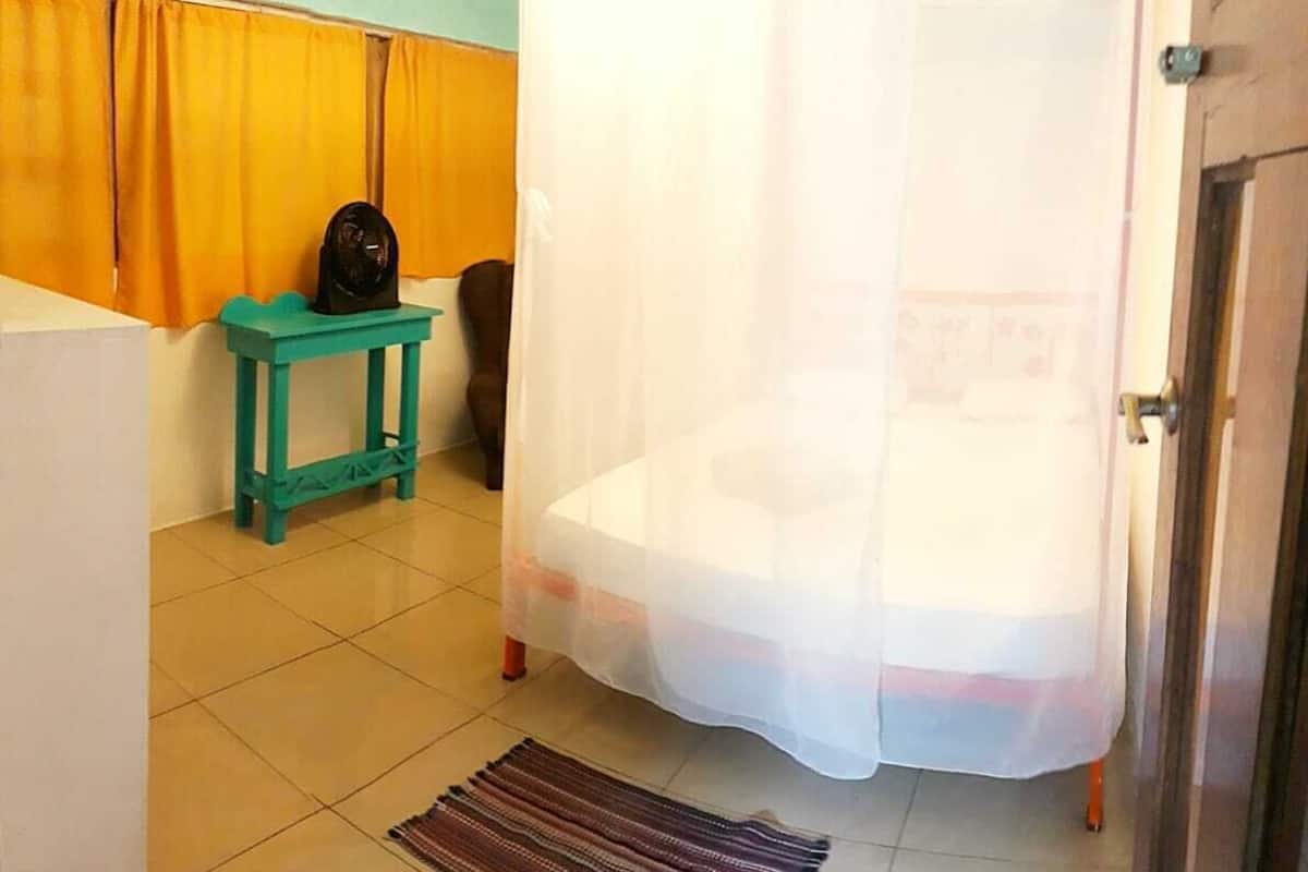 Quarto com uma cama, uma mesinha com um ventilador, uma cortina e uma porta.