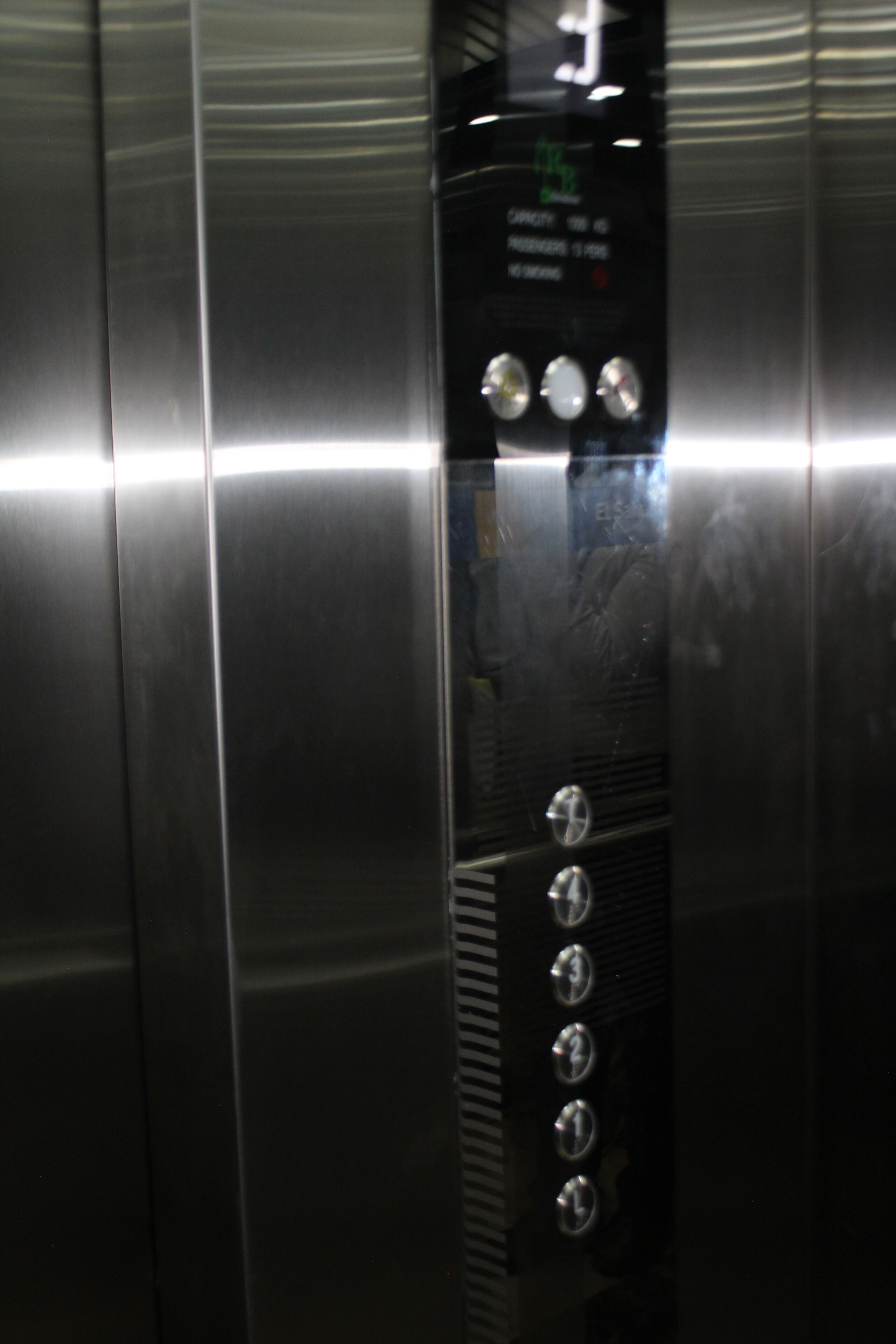 Interior de elevador com paredes metálicas e painel de comando com botões.