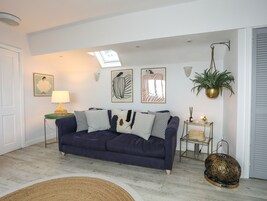 Uma sala de estar moderna com um sofá azul escuro, um tapete redondo e uma pequena mesa lateral com uma luminária. Há duas obras de arte emolduradas na parede, uma planta em vaso e um lustre.