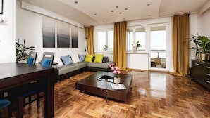 Uma sala de estar moderna com piso de madeira, uma mesa de centro escura, um sofá com almofadas, uma mesa de jantar com cadeiras e grandes janelas com cortinas.