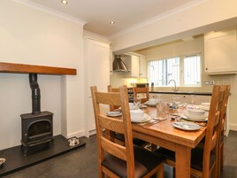Uma área de jantar com uma mesa de madeira posta para quatro pessoas, rodeada por cadeiras de madeira. Uma lareira a lenha preta está posicionada à esquerda da mesa.