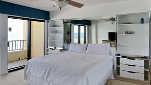 Quarto com cama grande, ventilador de teto, porta de correr para uma varanda e guarda-roupa branco com gavetas.