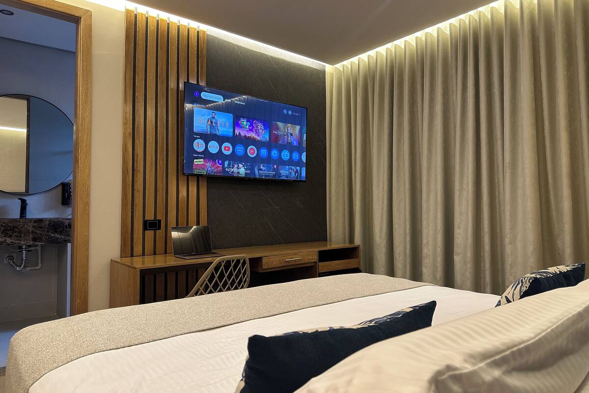 Quarto de hotel moderno com uma cama grande, televisão de tela plana na parede, mesa de console de madeira e uma grande janela com cortinas.