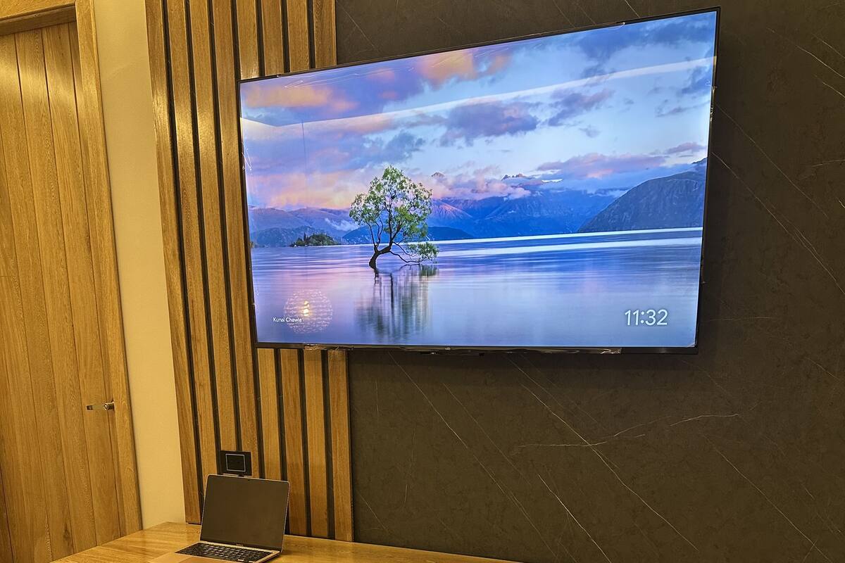 Quarto moderno com escrivaninha de madeira, um laptop, uma televisão exibindo uma paisagem cênica e uma cadeira.