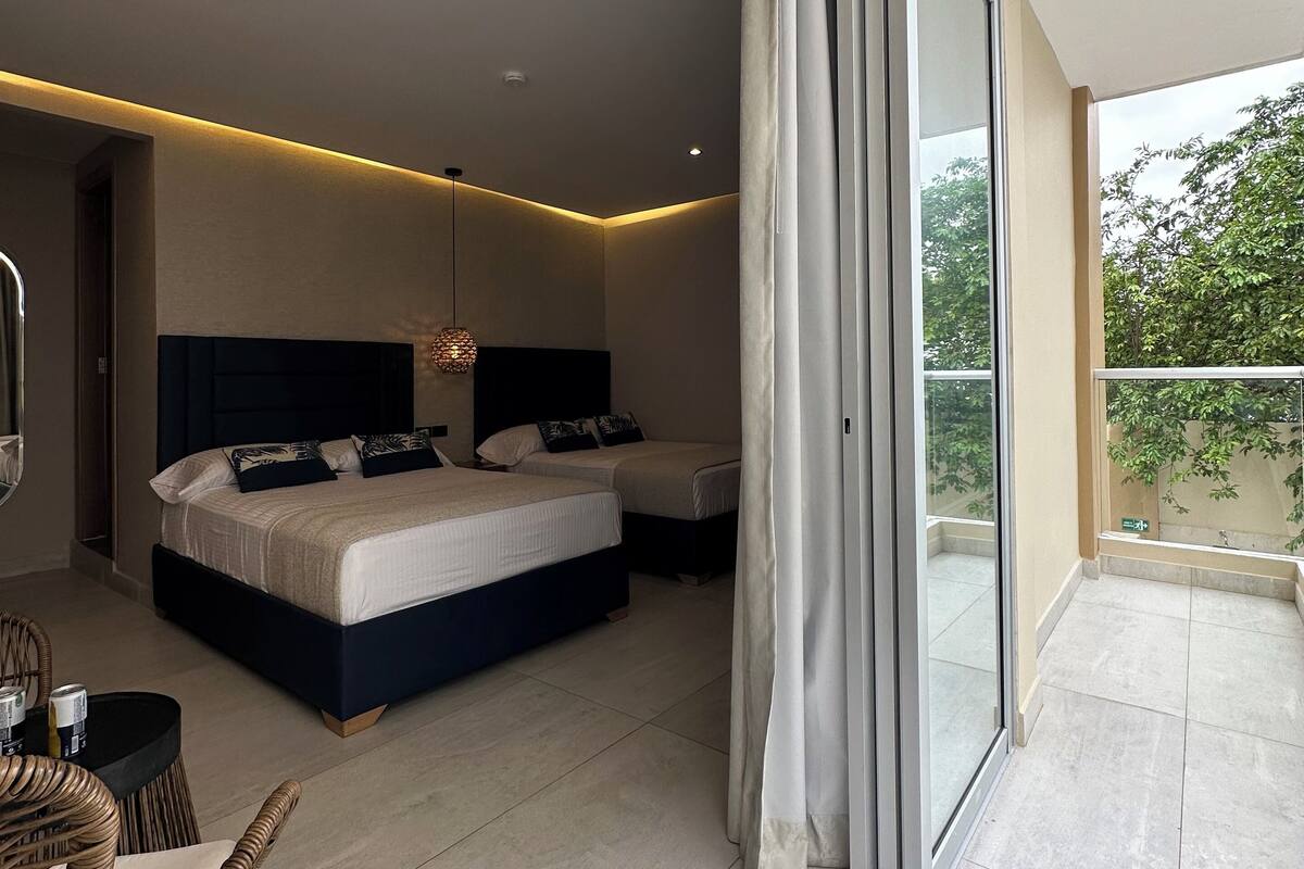 Quarto moderno com uma cama grande, duas camas menores, área de jantar com mesa e cadeiras, e varanda com vista para o verde.