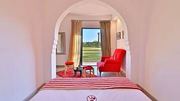 Quarto de hotel com uma cama grande, uma poltrona vermelha, uma mesa pequena e vista para uma paisagem verde através de uma janela com cortinas.