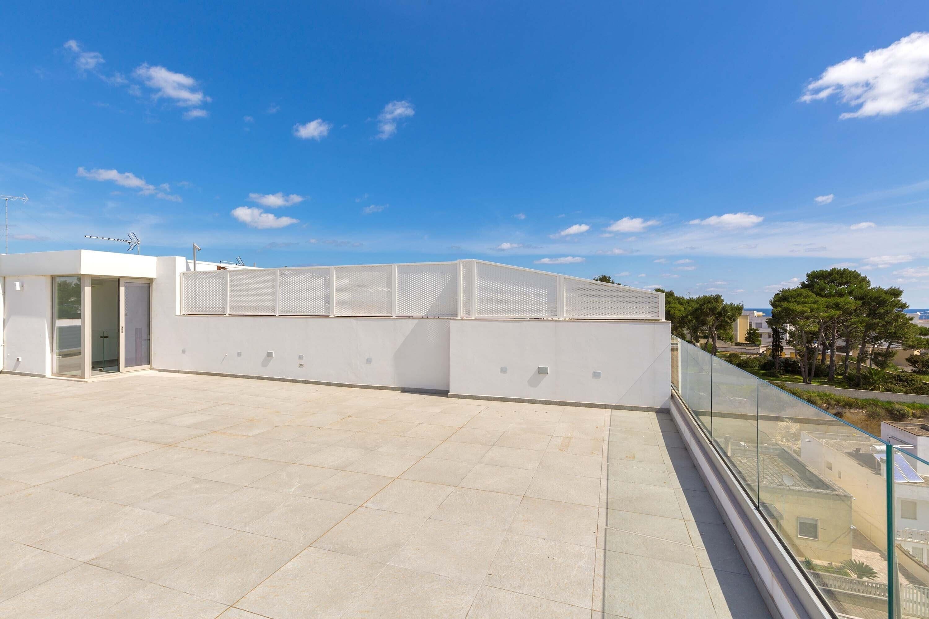 Terraço no telhado com guarda-corpo de vidro, um edifício branco e um céu azul límpido.