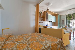 Quarto com cama, cadeira, mesinha e área de cozinha com armários e pia.