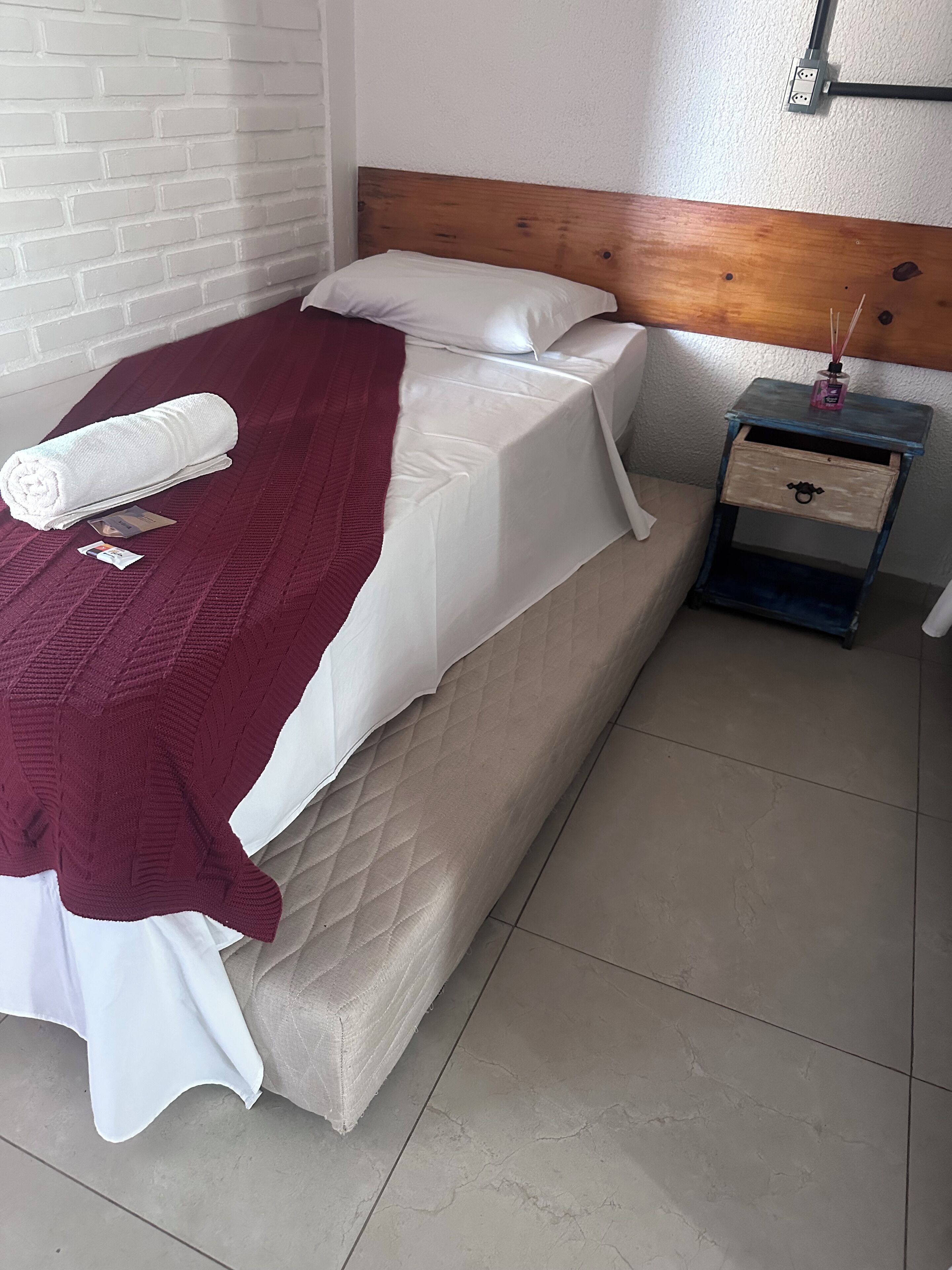 Uma cama bem arrumada com edredom branco e manta vermelha, cabeceira de madeira, mesa de cabeceira pequena com gaveta e uma tomada na parede.