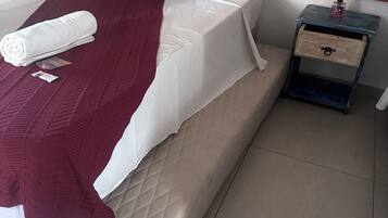Uma cama bem arrumada com edredom branco e manta vermelha, cabeceira de madeira, mesa de cabeceira pequena com gaveta e uma tomada na parede.