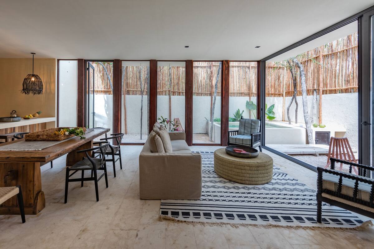 Cozinha moderna com mesa de madeira, cadeiras e luminárias pendentes. Há uma área de estar com sofá e um banquinho, e vista para o exterior através de grandes janelas.