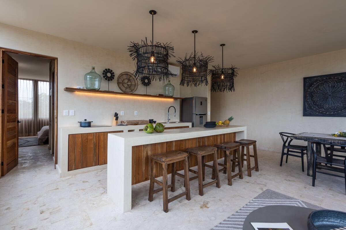 Uma cozinha moderna com ilha central, banquetas de madeira, luminárias pendentes e um espaço de refeições com mesa e cadeiras.
