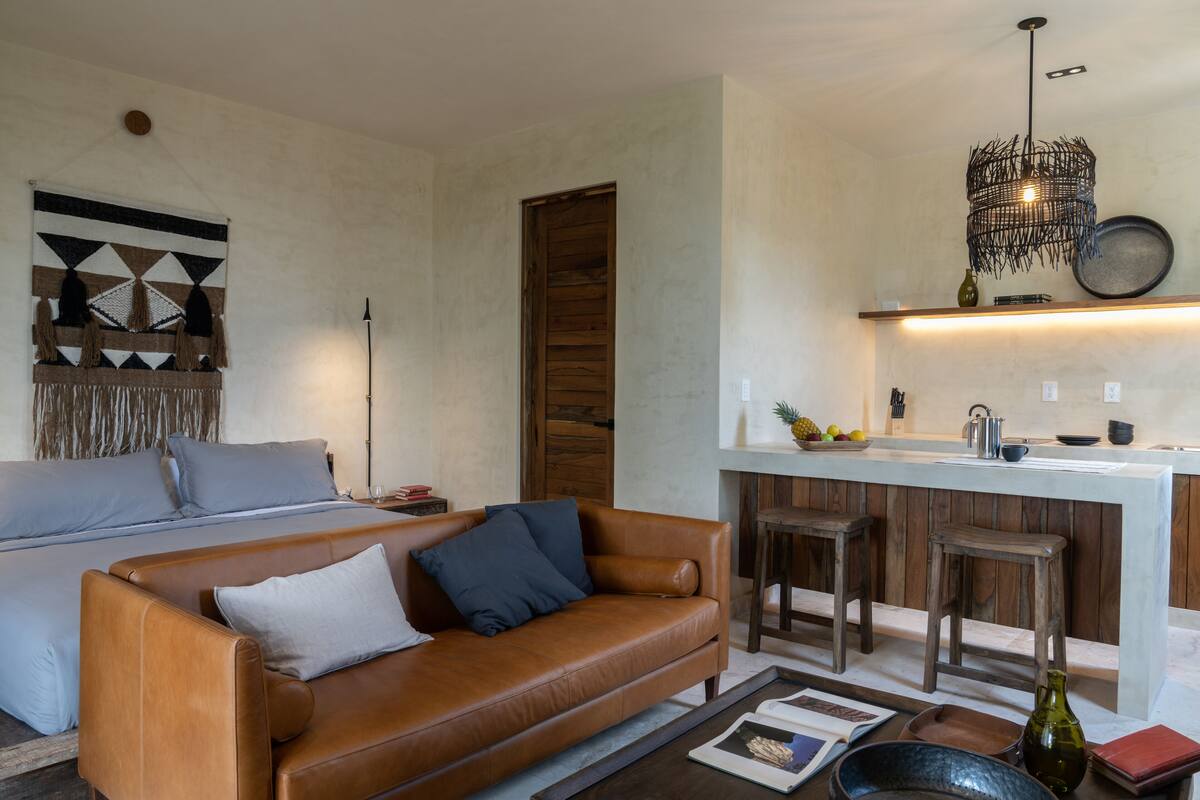 Uma sala moderna com um sofá de couro marrom, uma porta de madeira e uma área de cozinha com bancada branca e bancos de madeira.