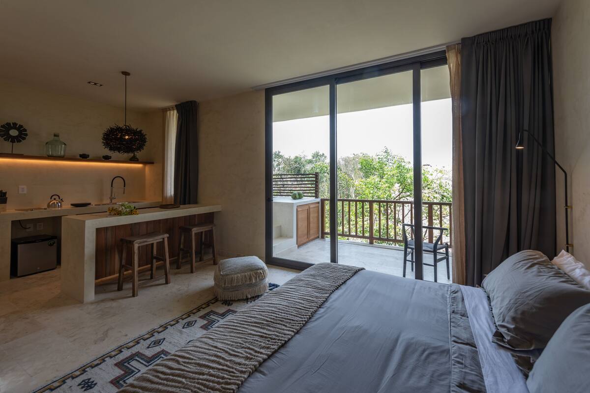 Quarto moderno com cama grande, pequeno espaço de cozinha e varanda com vista para o verde.