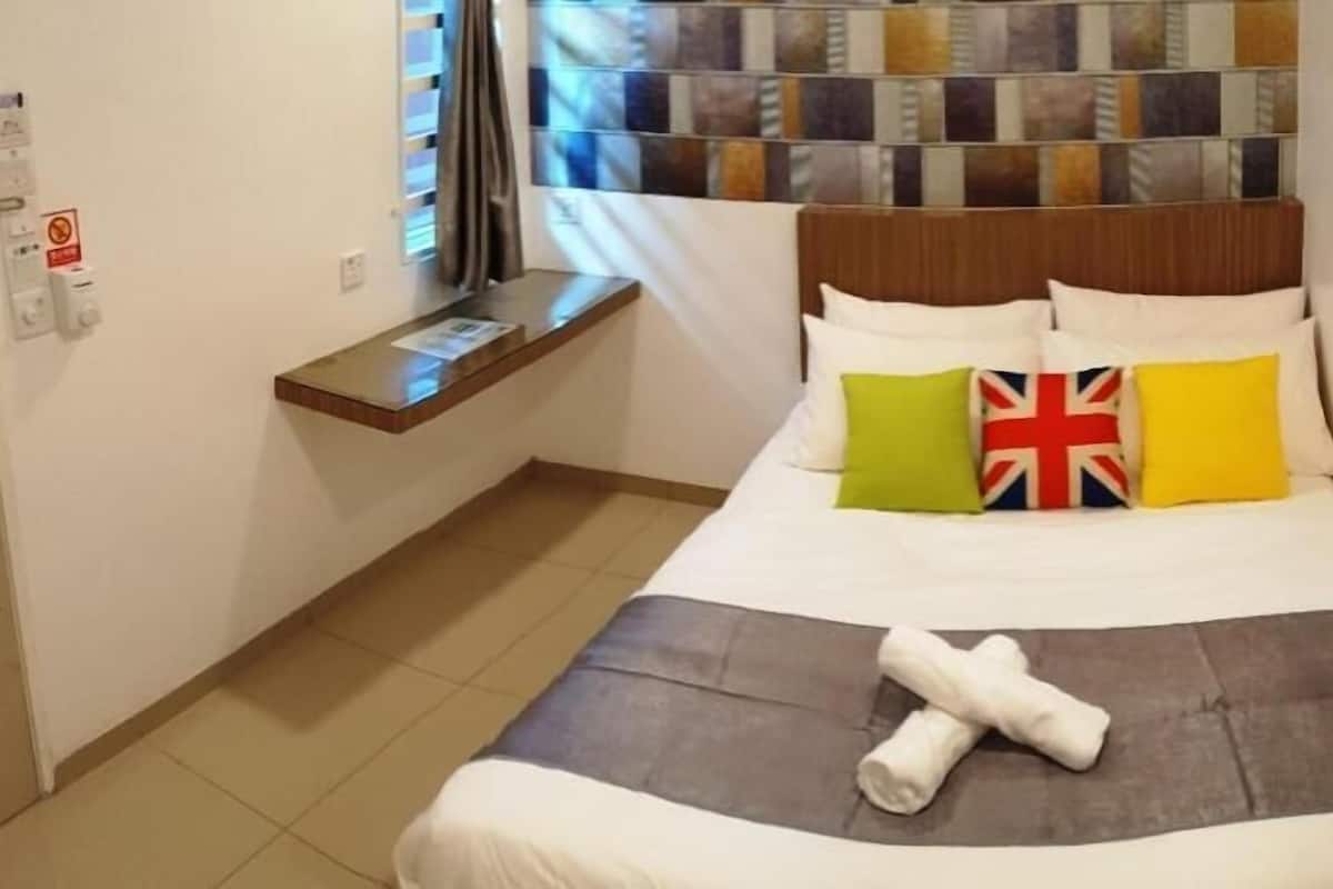 Quarto de hotel com uma cama, uma janela com persianas, uma pequena prateleira e um detector de incêndio fixado na parede.