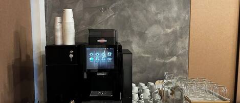 Máquina de café com visor digital, xícaras empilhadas e uma bandeja com copos e copos descartáveis sobre uma mesa de madeira.