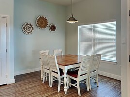 Uma sala de jantar com mesa de madeira e cadeiras brancas, três decorações na parede e uma janela com persianas.